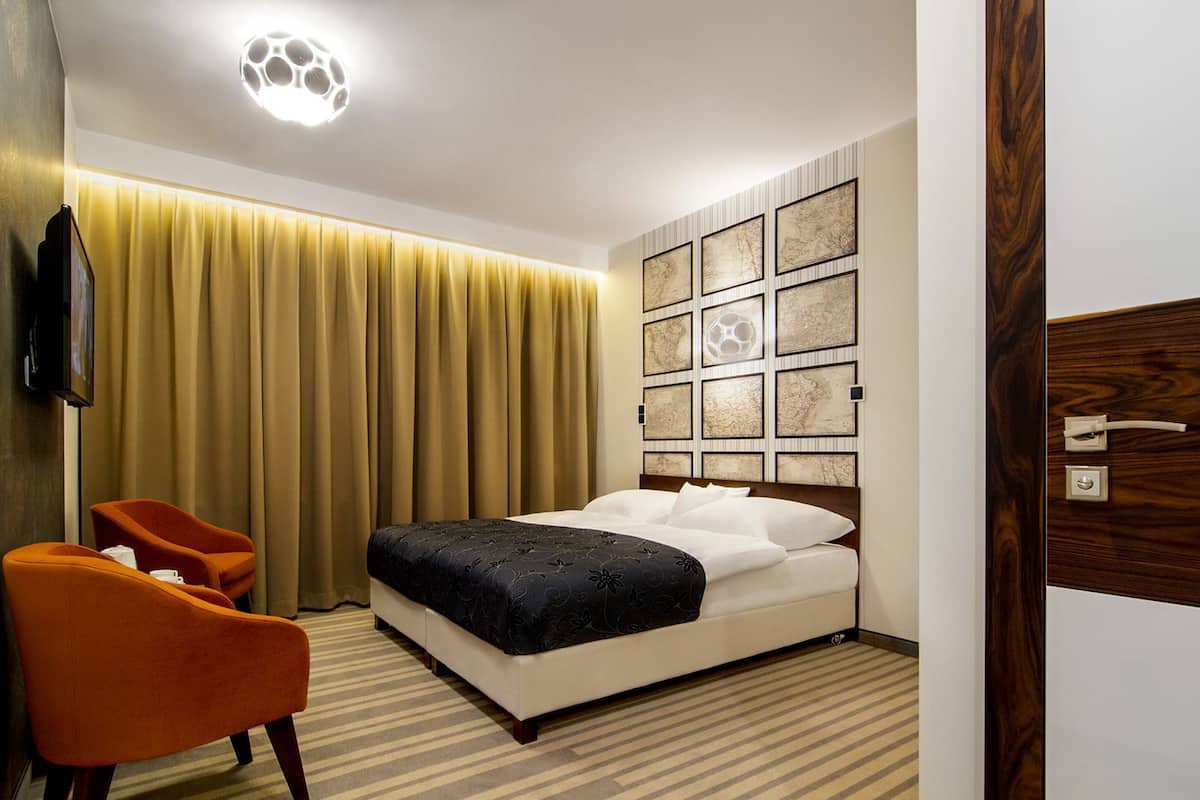 Quarto de hotel com uma cama, televisão, cadeira e parede com obras de arte emolduradas.