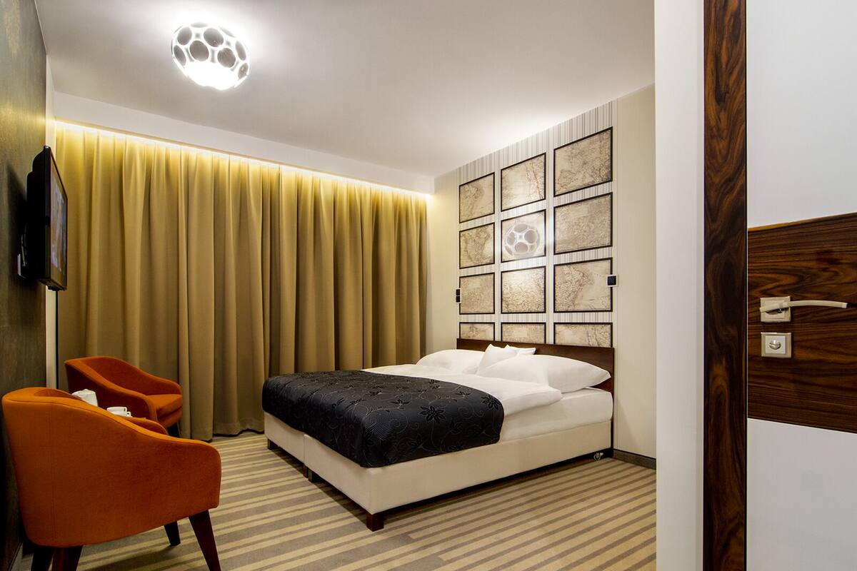 Quarto de hotel com cama, televisão, área de estar com cadeiras e parede com obras de arte emolduradas.