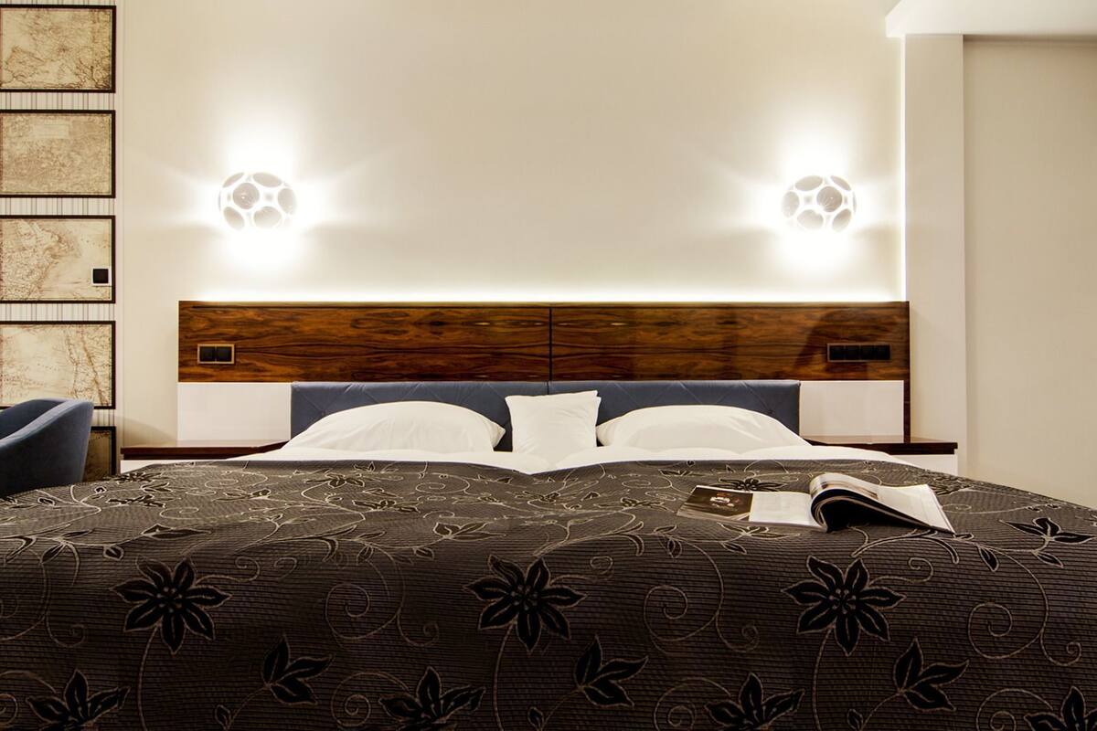 Uma cama bem arrumada com colcha estampada escura, cabeceira de madeira e duas luminárias fixadas na parede.