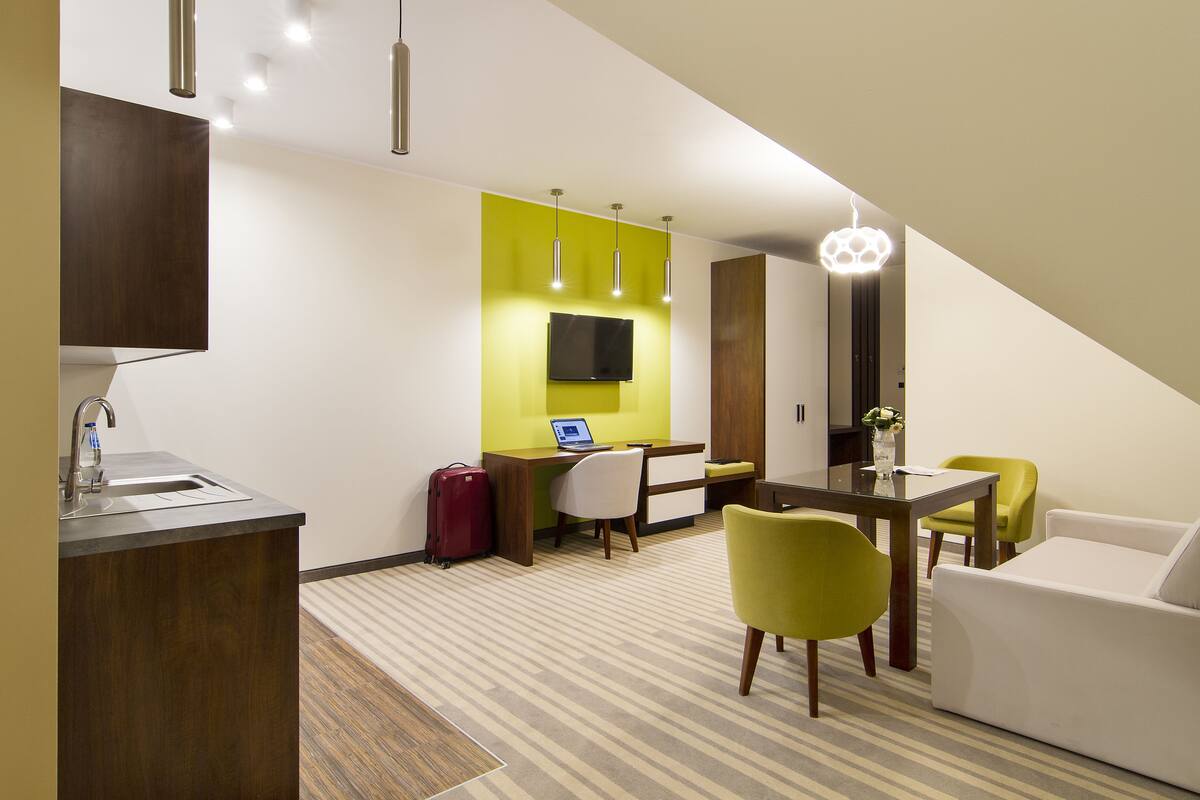 Quarto de hotel moderno com cozinha compacta, uma escrivaninha com computador, área de refeições com cadeiras e uma TV fixada na parede.
