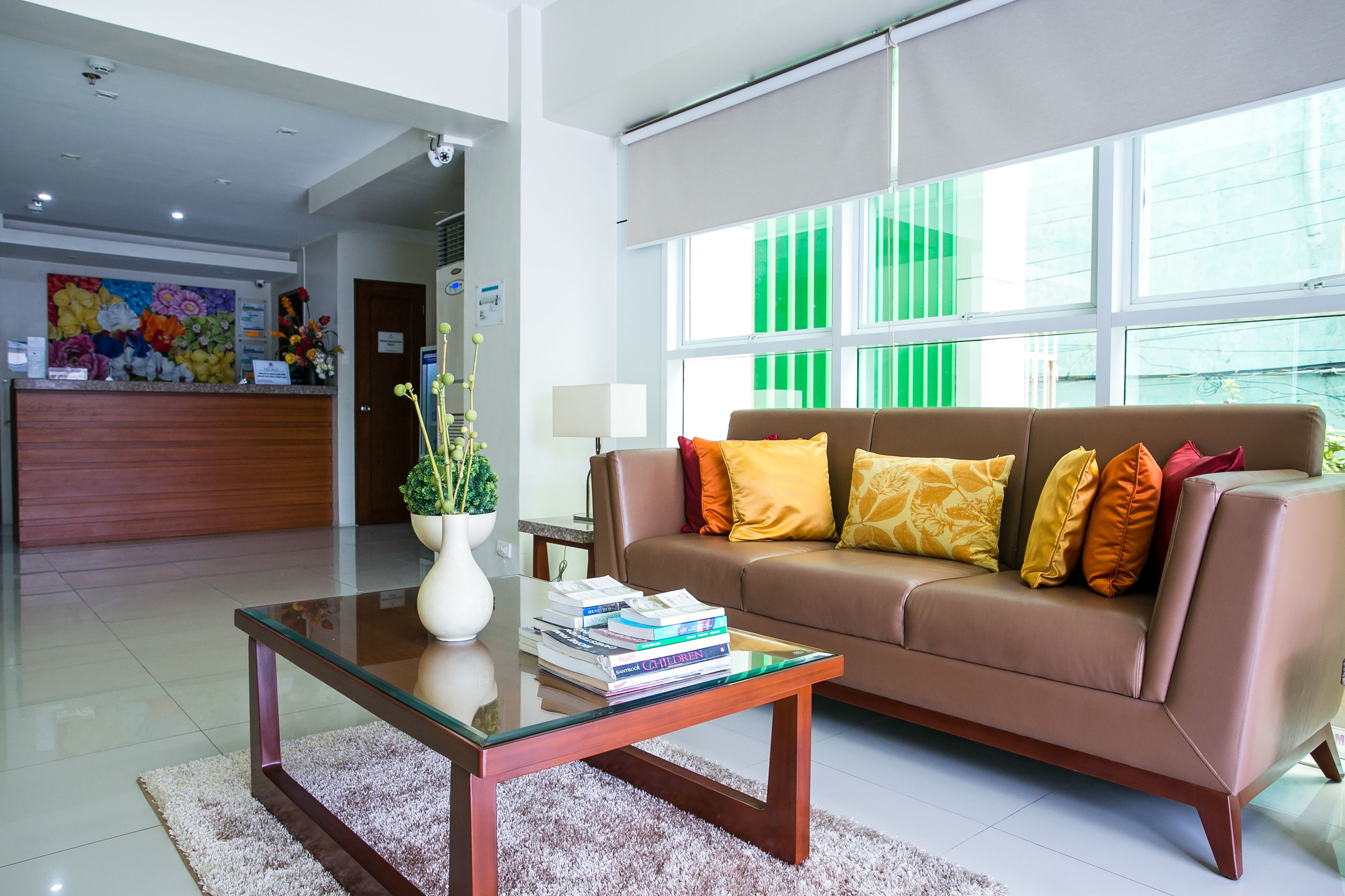 Um lobby de hotel moderno com um balcão de recepção, um sofá de couro marrom com almofadas coloridas, uma mesa de centro de vidro com livros e uma janela grande com persianas verdes.