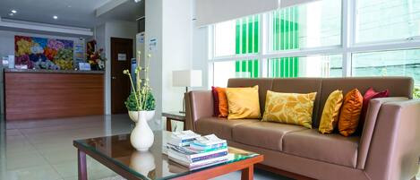 Um lobby de hotel moderno com um balcão de recepção, um sofá de couro marrom com almofadas coloridas, uma mesa de centro de vidro com livros e uma janela grande com persianas verdes.