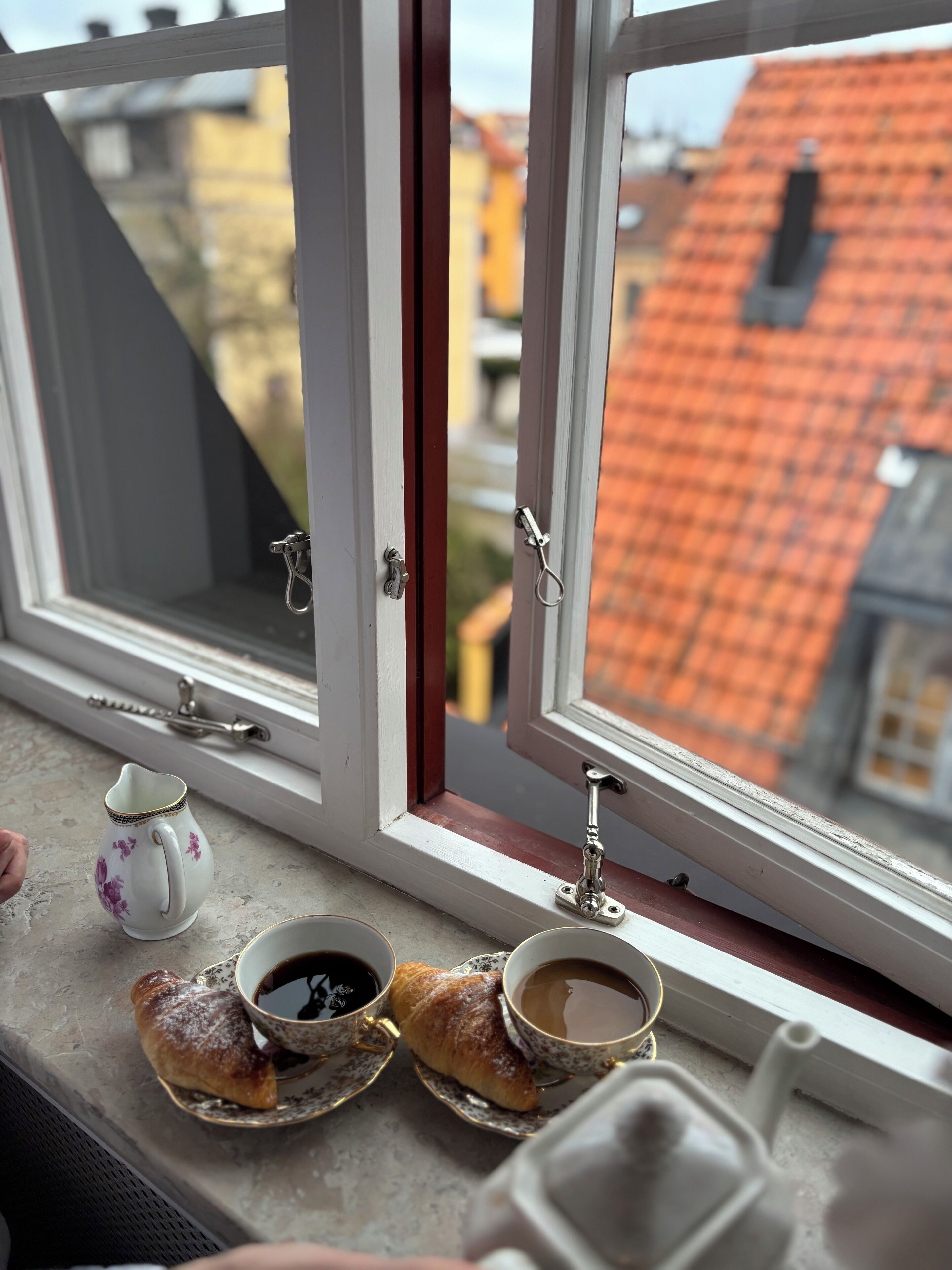 Uma janela com vista para edifícios, uma chávena de café, um croissant e um bule na soleira.