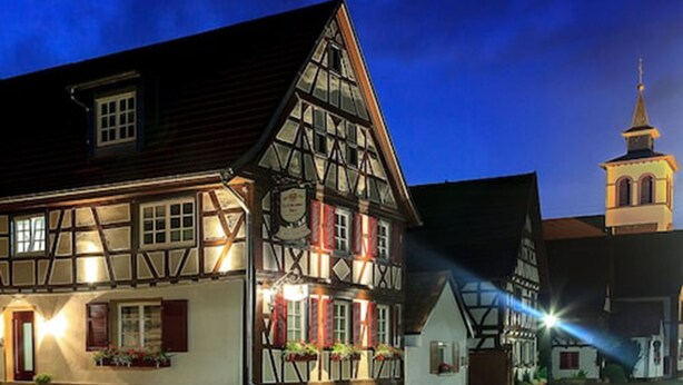 Um edifício tradicional em estilo de enxaimel com um relógio, janelas iluminadas e o topo de uma igreja ao fundo.
