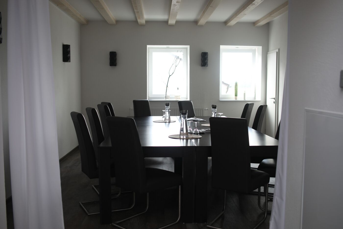 Uma sala de conferências com uma mesa retangular, cadeiras pretas e duas janelas que proporcionam iluminação natural.