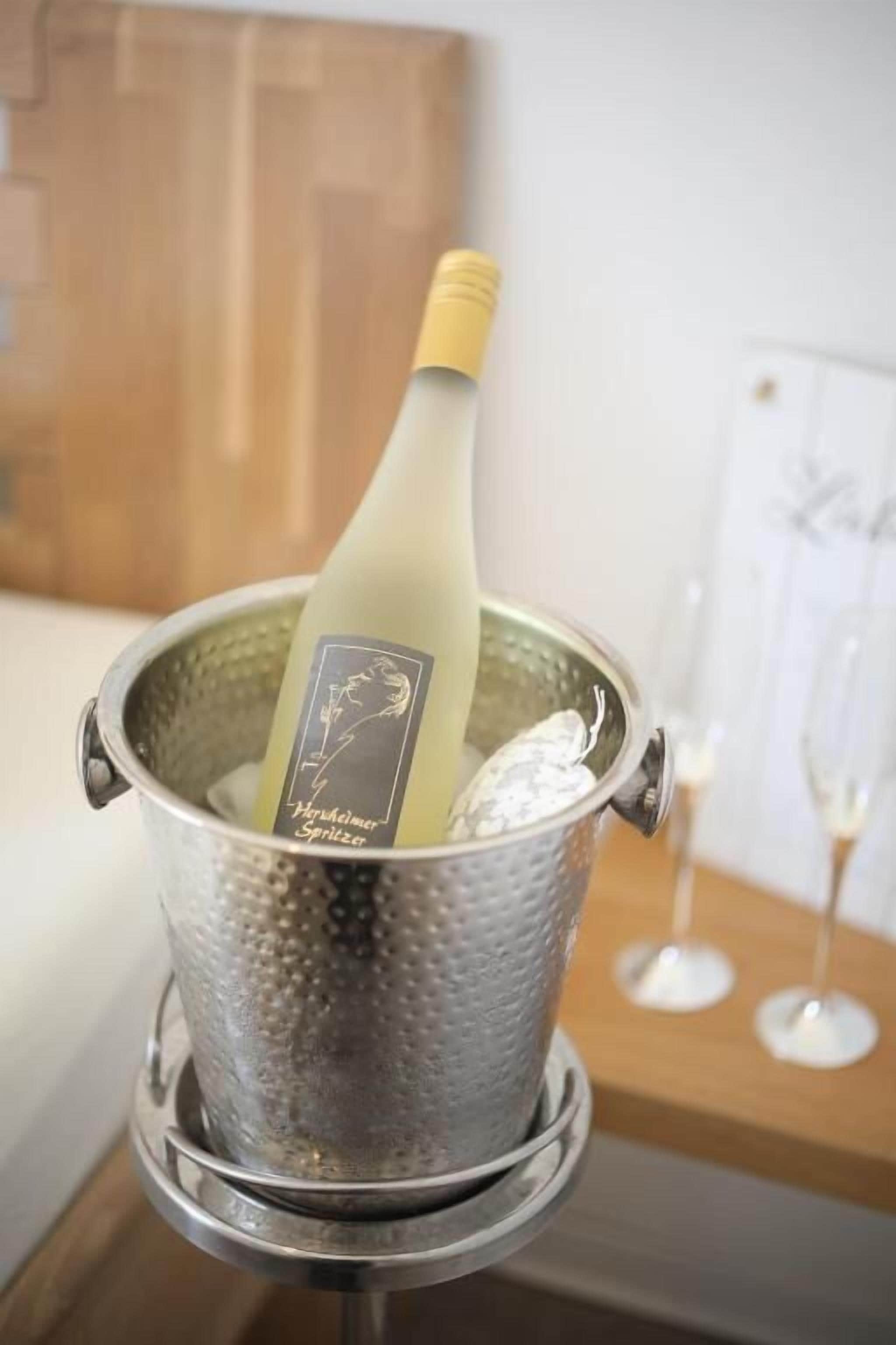 Uma garrafa de vinho em recipiente metálico com etiqueta, colocada sobre uma mesa com um copo.