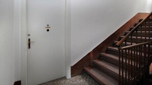 Uma porta branca com puxador de latão e fechadura, dando acesso a uma escada de madeira com corrimão de metal.