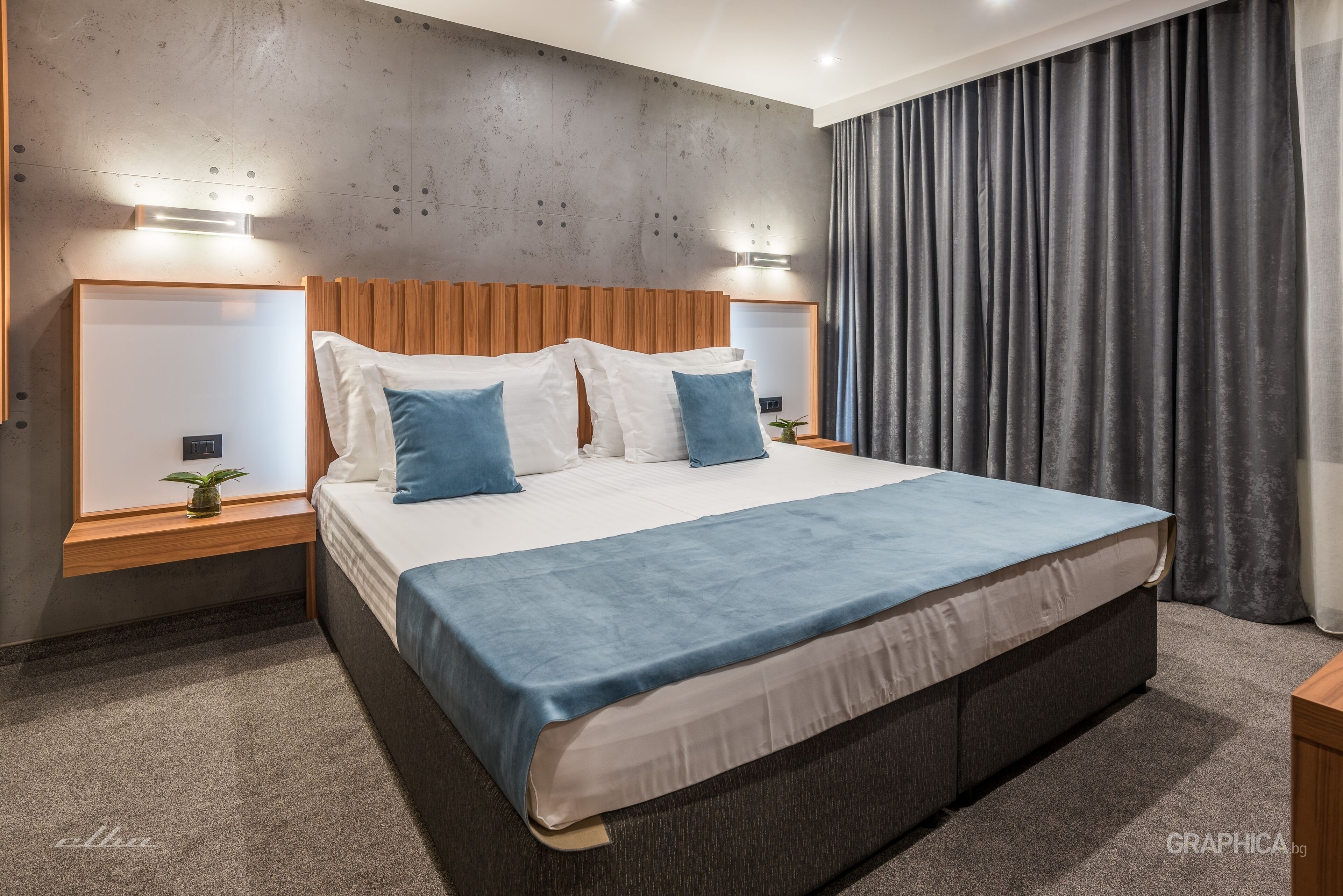Quarto de hotel moderno com uma cama grande, cabeceira de madeira, mesas de cabeceira com abajures e uma cortina cinza.