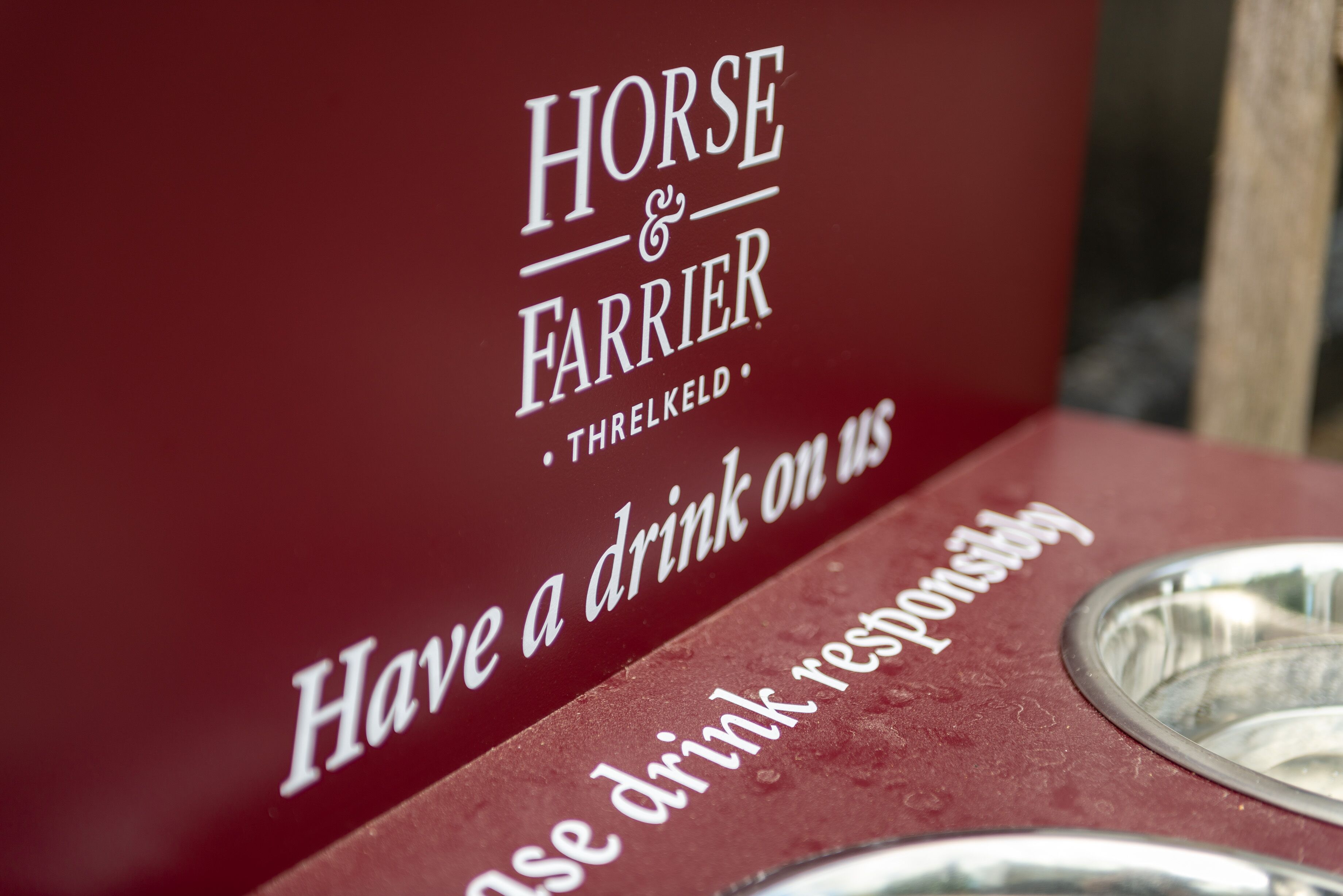 Uma placa vermelha com os dizeres 'HORSE & FARRIER' e 'Bebida por nossa conta', indicando um bar ou restaurante.