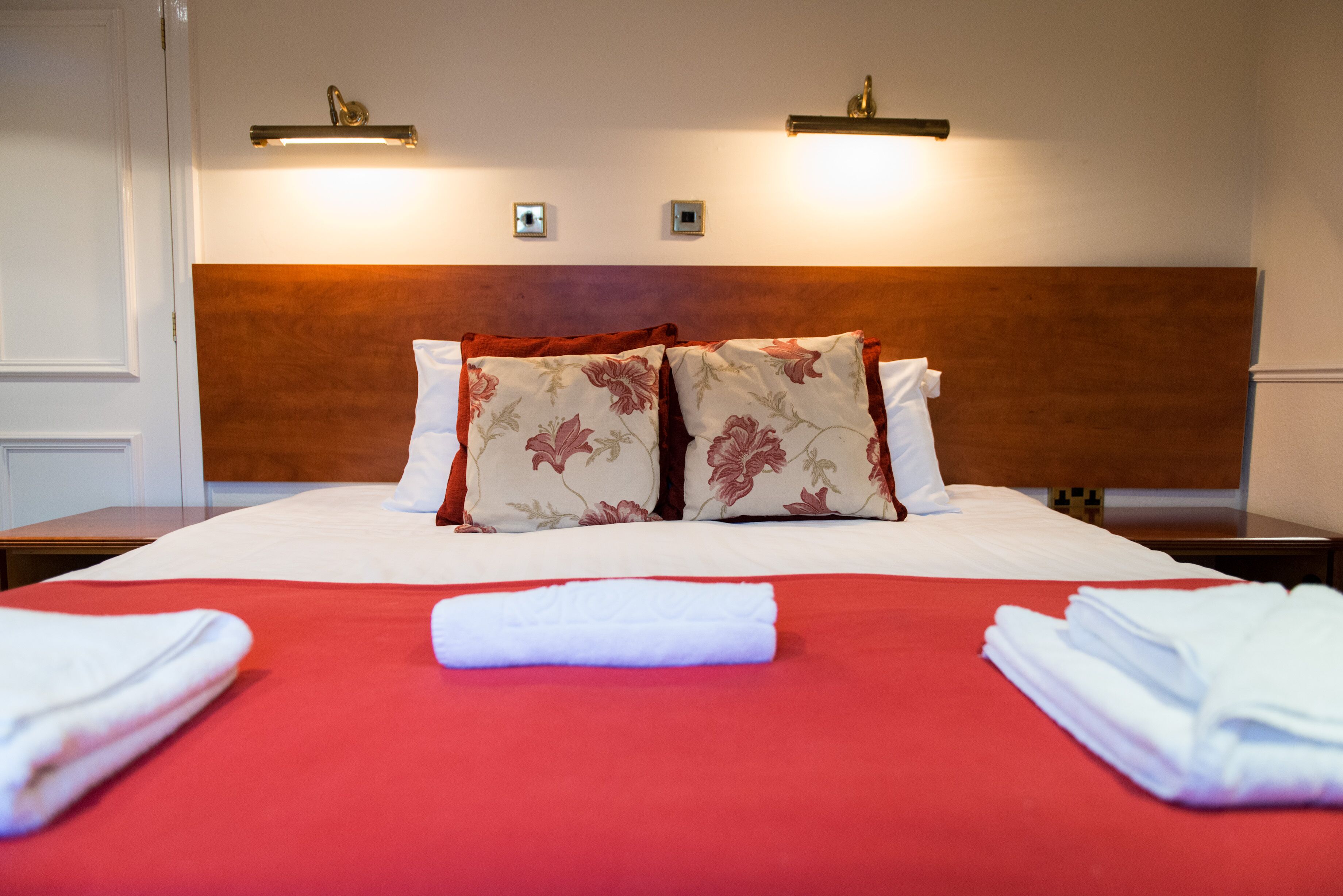 Cama bem arrumada com almofadas florais, colcha vermelha e toalhas brancas. Há duas mesas de cabeceira com luminárias e uma cabeceira de madeira.