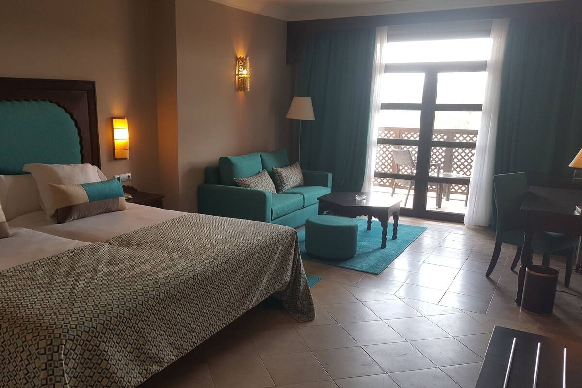 Quarto de hotel com duas camas, um sofá, uma mesinha e vista para o exterior.