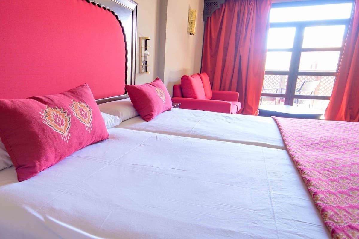 Quarto de hotel com duas camas, cada uma com lençóis brancos e almofadas vermelhas. Há uma cabeceira vermelha, uma janela com cortinas vermelhas e vista para móveis de exterior.