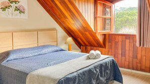 Quarto com teto inclinado, paredes de madeira, cama com colcha azul e branca, mesa de cabeceira com abajur e janela com vista para vegetação.