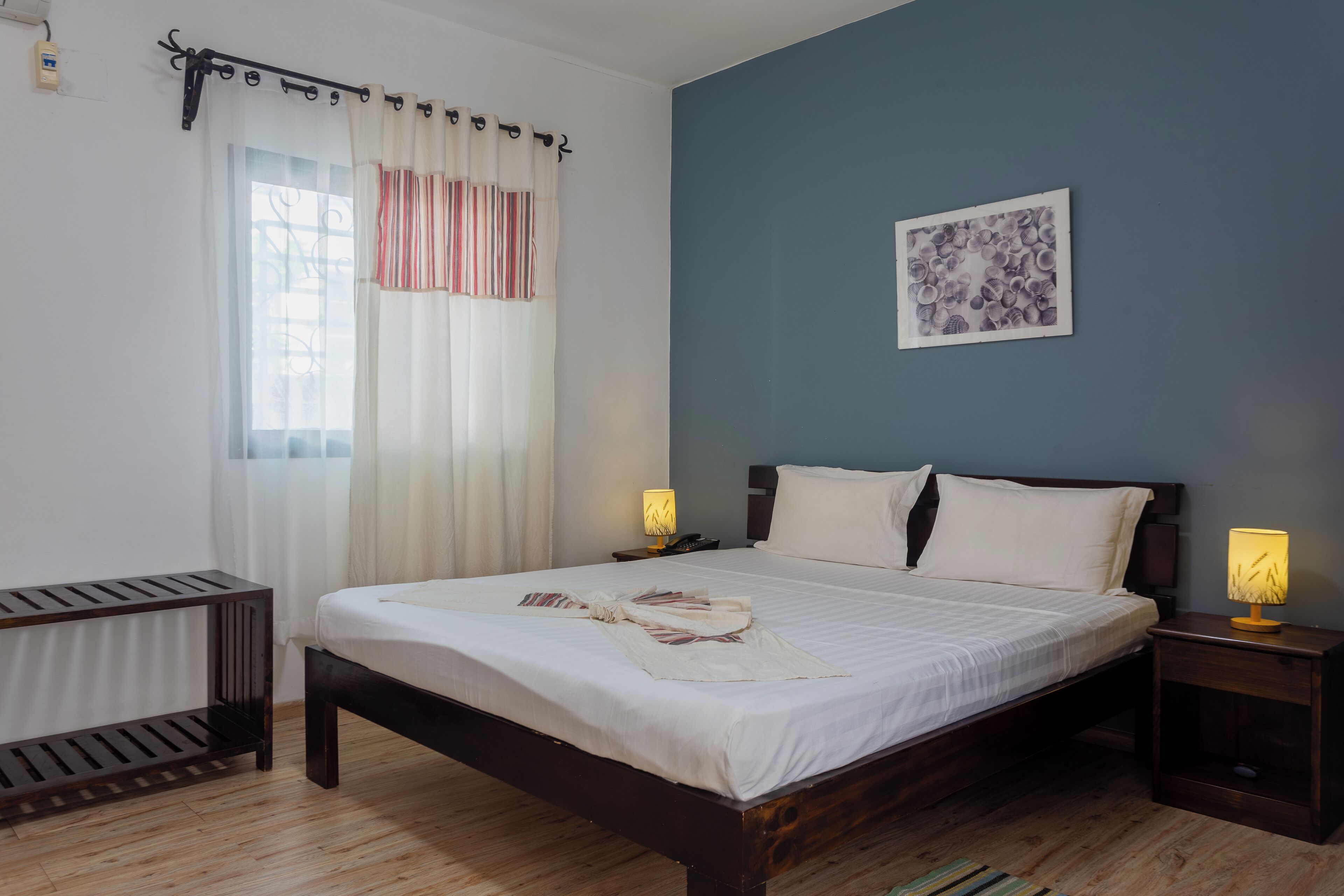 Quarto com cama de madeira, roupa de cama branca, mesa de cabeceira com abajur, janela com cortinas e quadro pendurado na parede.