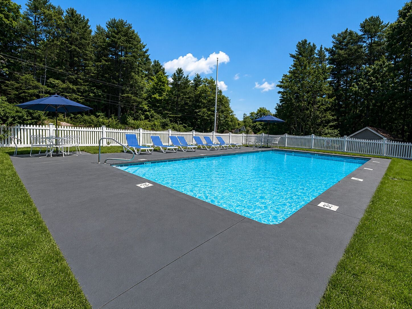 Piscina retangular com água azul, rodeada por uma cerca branca e equipada com espreguiçadeiras e guarda-sóis.