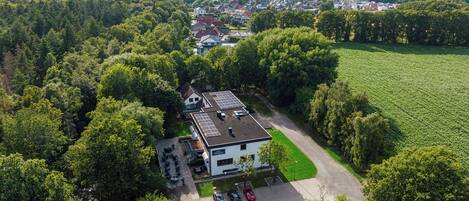 Vista aérea de um bairro residencial com uma casa moderna de dois andares, painéis solares, vegetação exuberante e uma pequena área de estacionamento.