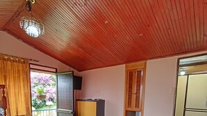 Quarto com teto de madeira, cama com lençóis brancos, guarda-roupa de madeira, mesa pequena com uma cadeira e janela com cortinas.