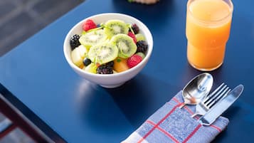 Uma mesa com uma tigela de salada de frutas, um copo de suco de laranja e talheres sobre uma toalha de mesa azul.