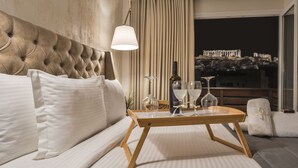 Quarto de hotel com cabeceira estofada, mesinha de madeira com copos e uma garrafa, e vista para a paisagem urbana pela janela.