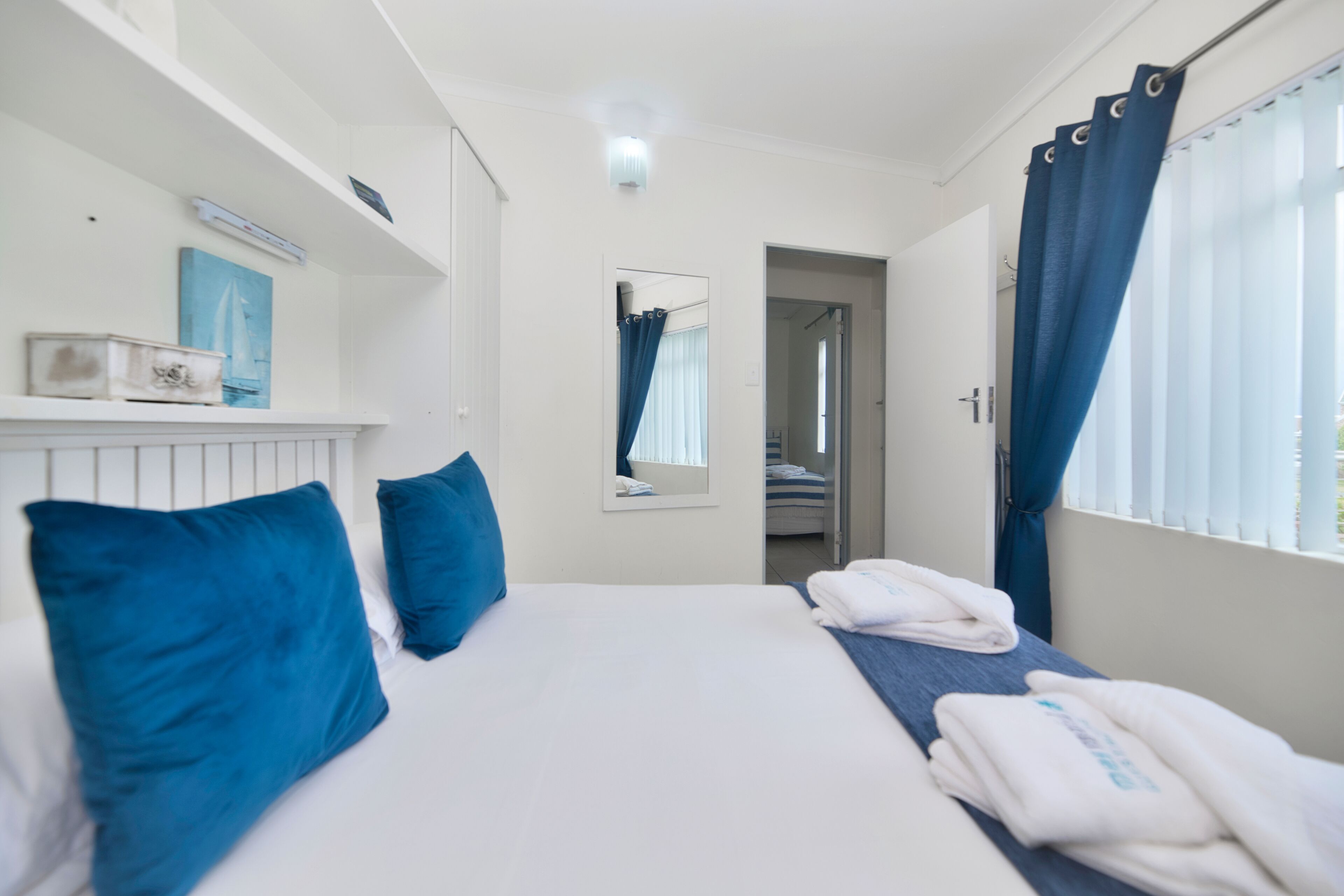 Quarto com cama, duas almofadas, janela com cortinas, espelho e prateleira com um quadro e um objeto decorativo.