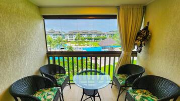 Varanda com cadeiras de vime e uma mesa de vidro, com vista para uma piscina e edifícios do resort.