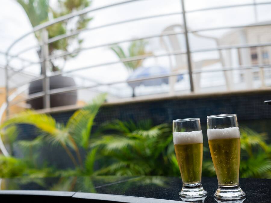 Duas cervejas sobre uma superfície refletora, com um fundo desfocado de vegetação e um edifício.