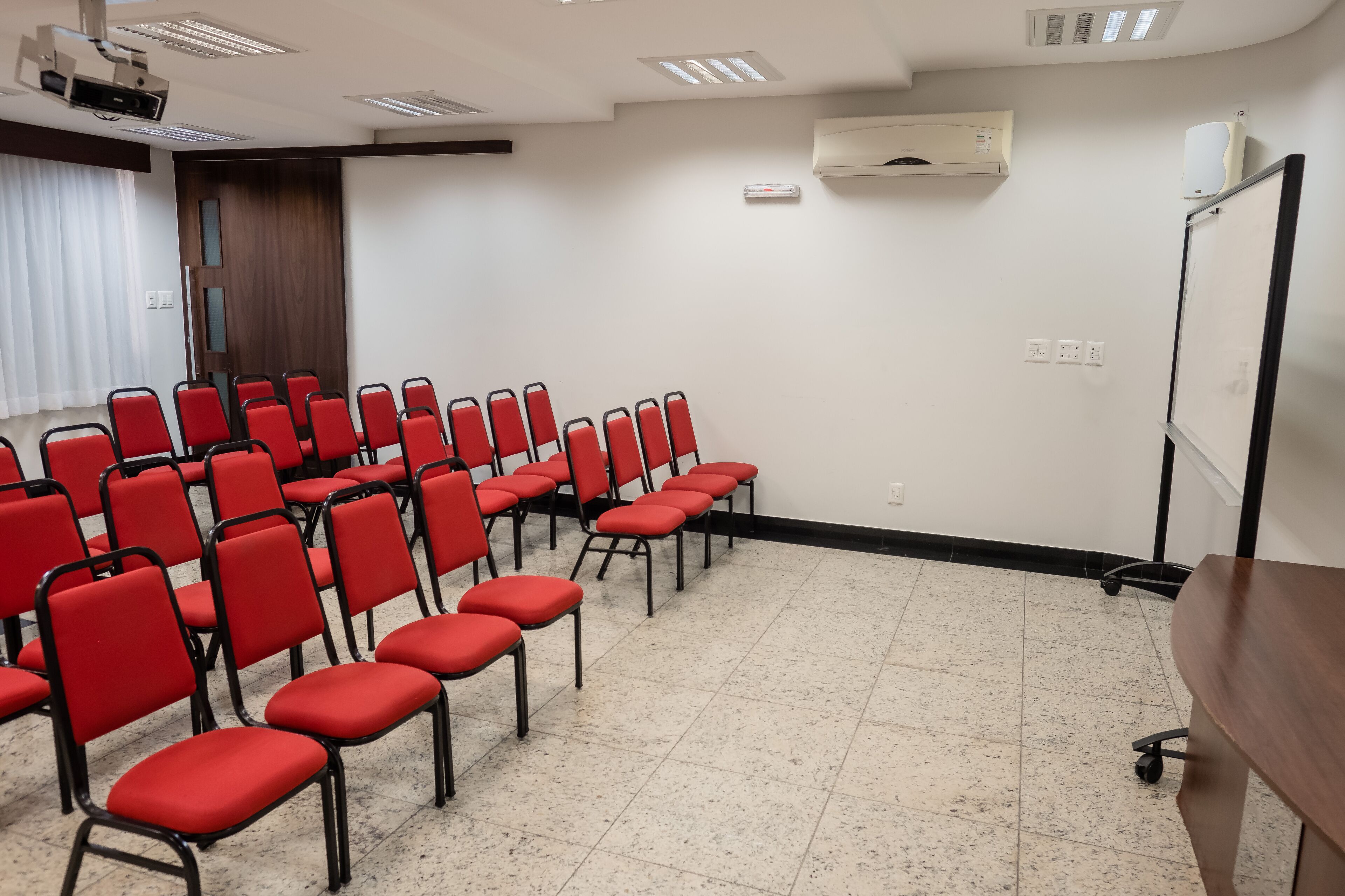 Sala de conferências com fileiras de cadeiras vermelhas, um projetor, um aparelho de ar condicionado e um quadro branco.