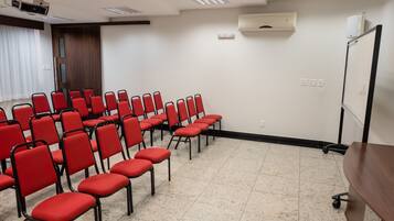 Sala de conferências com fileiras de cadeiras vermelhas, um projetor, um aparelho de ar condicionado e um quadro branco.