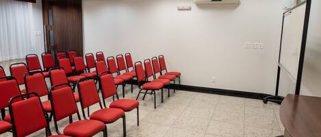 Sala de conferências com fileiras de cadeiras vermelhas, um projetor, um aparelho de ar condicionado e um quadro branco.
