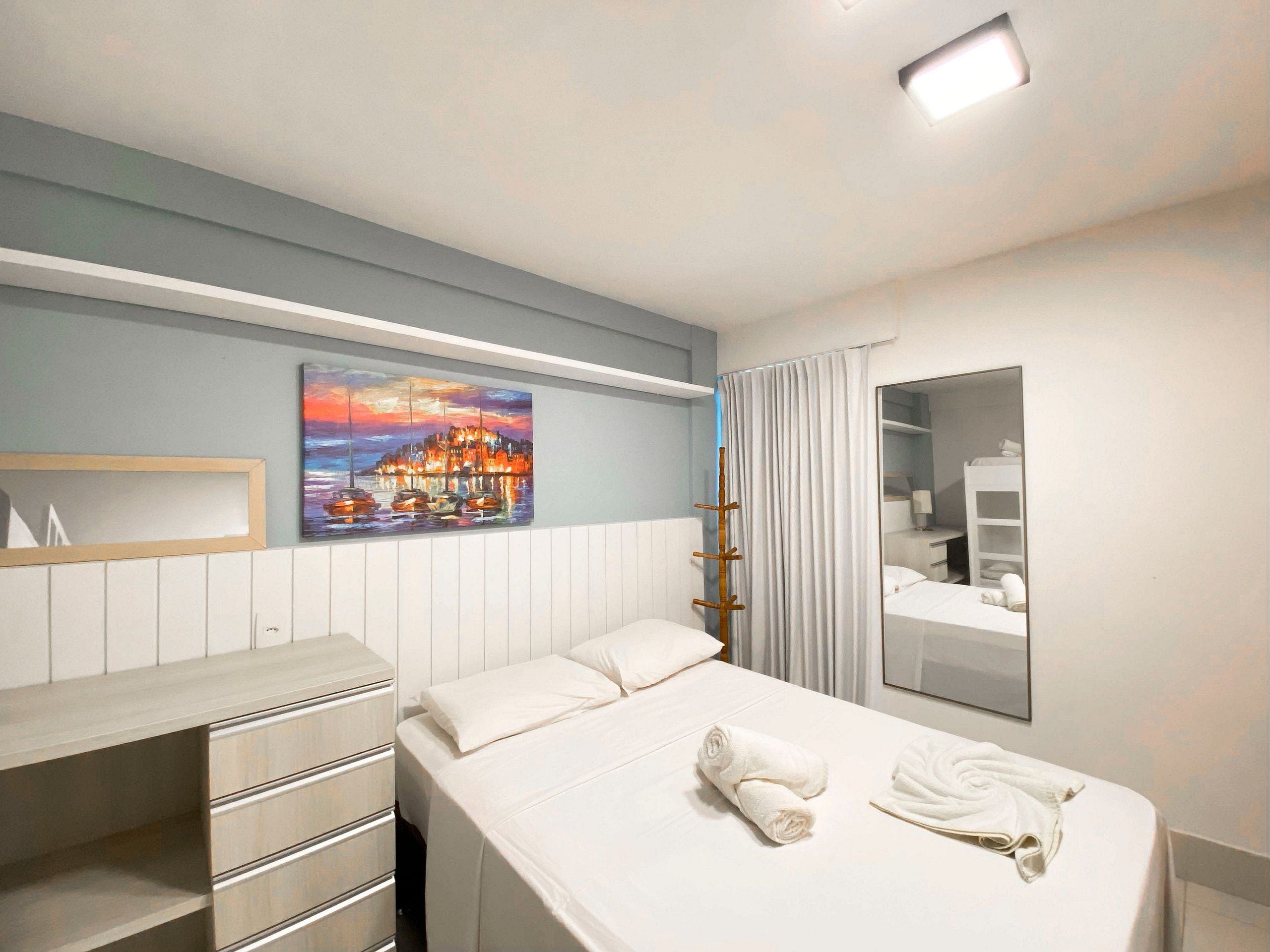 Quarto de hotel com uma cama, penteadeira, quadro, espelho e janela com cortinas.