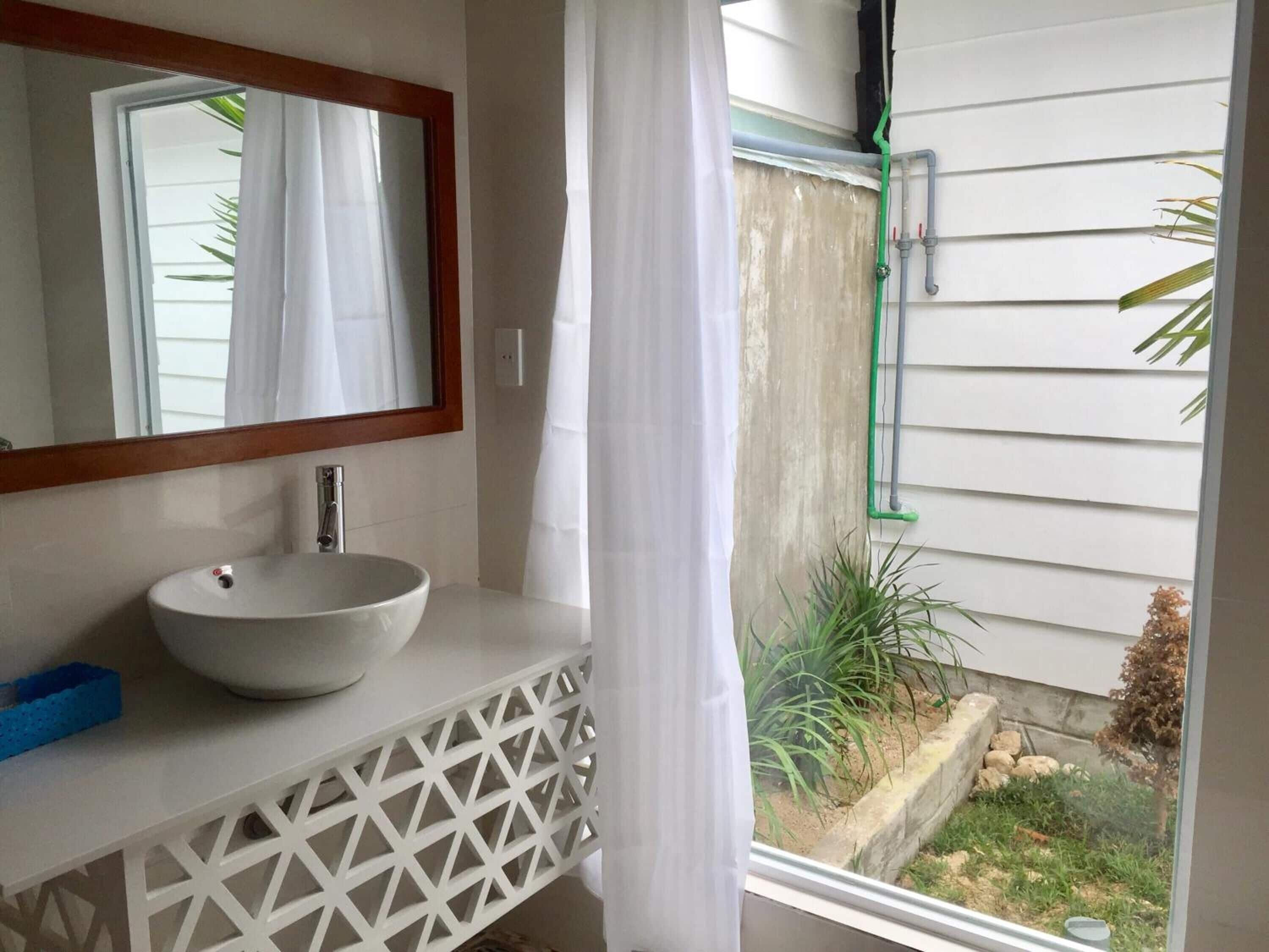 Um banheiro com pia branca, espelho e janela com vista para um jardim.