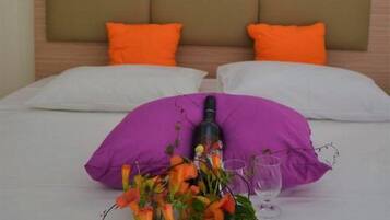 Quarto de hotel com uma cama, dois travesseiros laranja e um roxo, e um arranjo de flores sobre a cama.