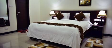 Cama bem arrumada com lençóis brancos e duas almofadas castanhas, mesa de cabeceira com abajur, quadro na parede e porta à esquerda.