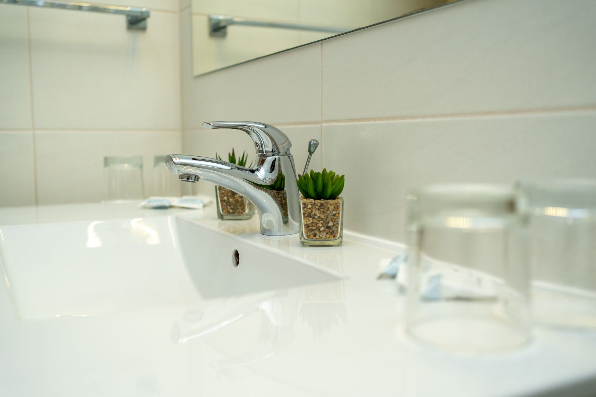 Casa de banho com lavatório branco, torneira cromada e duas pequenas plantas em vasos sobre o balcão.