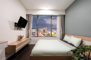 Quarto de hotel com cama, televisão na parede, janela com cortinas e vista noturna da cidade.