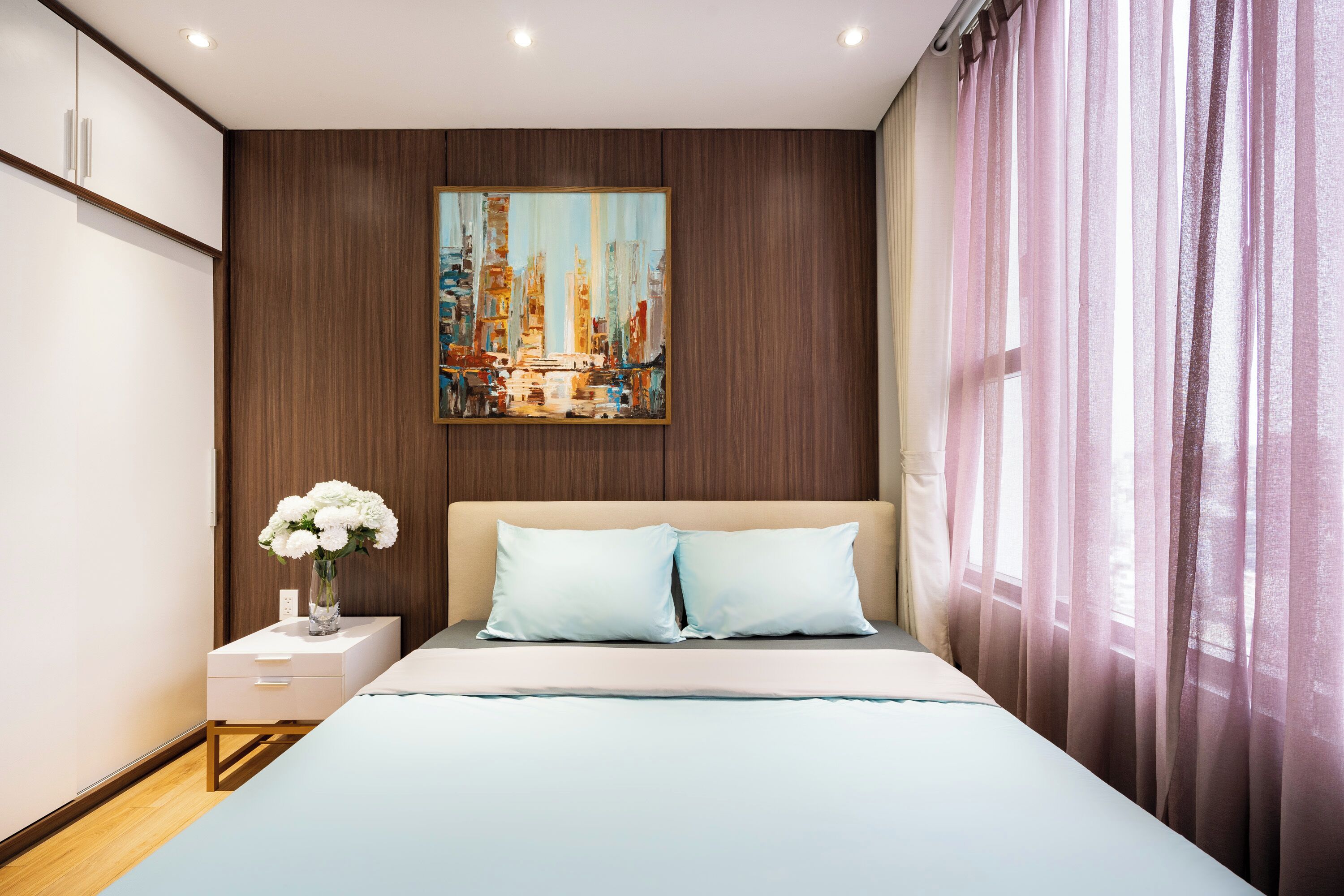 Quarto com cama, mesa de cabeceira, um vaso de flores e um quadro na parede.