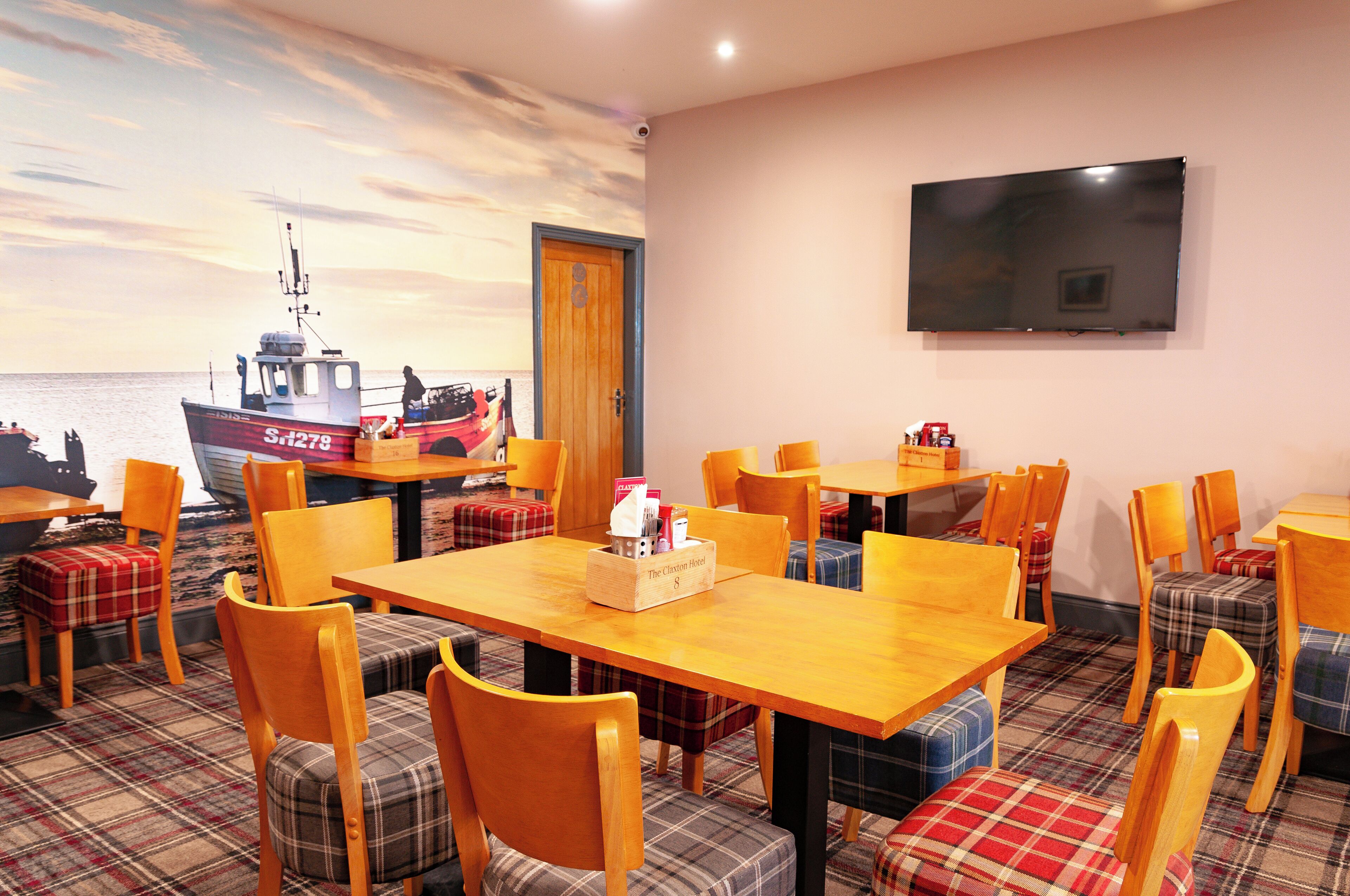 Um restaurante com mesas e cadeiras de madeira, uma televisão na parede e um mural de um barco.