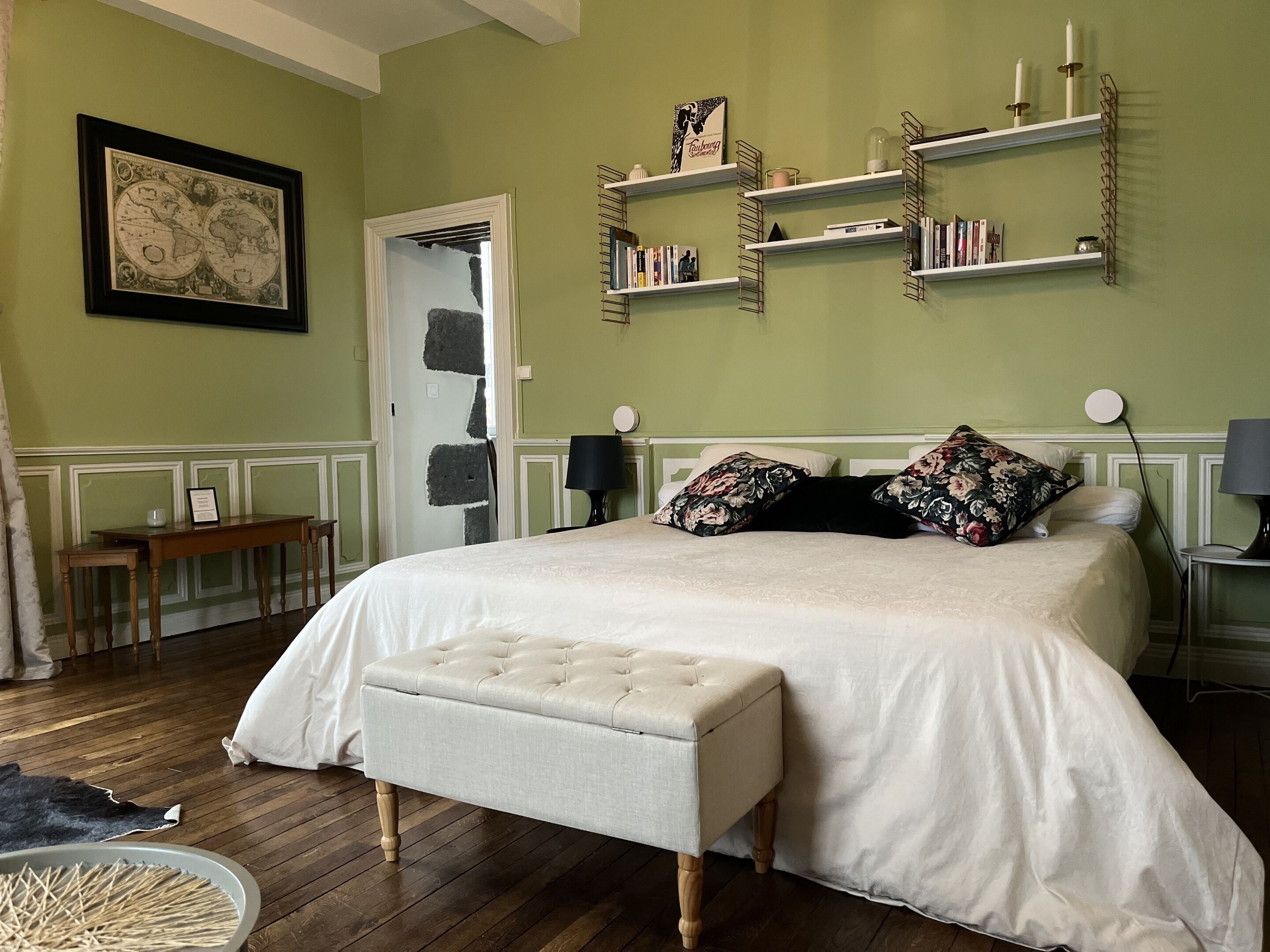 Quarto com cama, um banquinho, escrivaninha e prateleiras com livros e objetos decorativos.