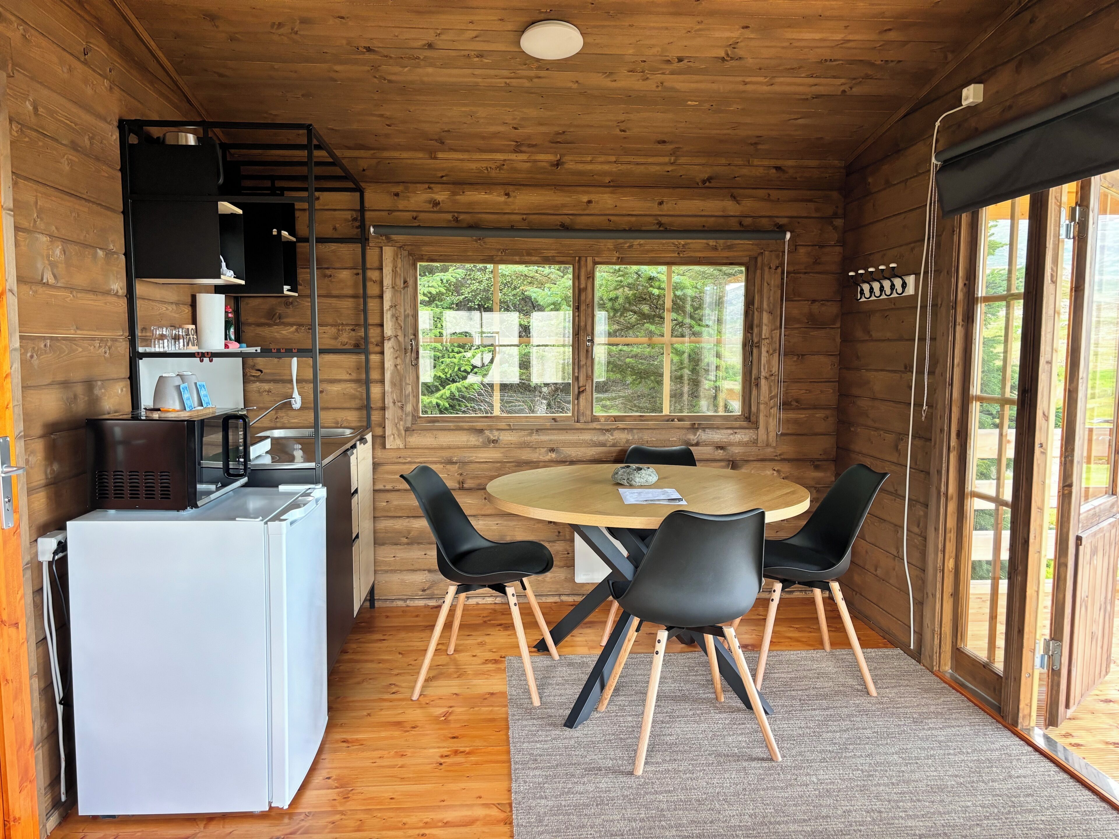 Cozinha compacta com geladeira, micro-ondas e cafeteira. Há uma mesa de jantar com quatro cadeiras e uma janela com vista para o verde lá fora.