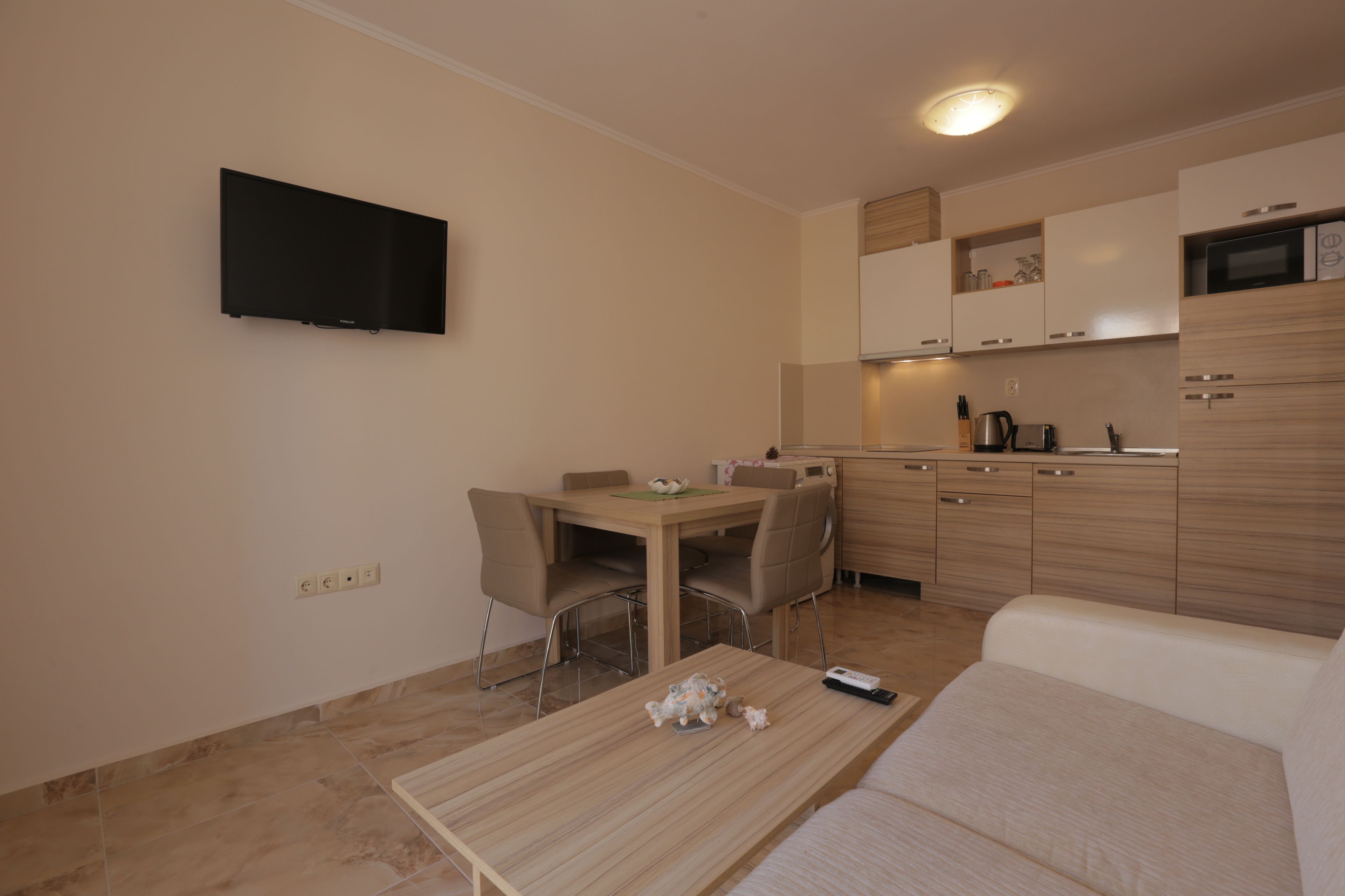 Um espaço de estar compacto com televisão de ecrã plano na parede, mesa de jantar com cadeiras, sofá e área de kitchenette com armários e micro-ondas.