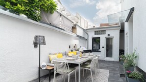 Uma área moderna de refeições ao ar livre, com mesa e cadeiras brancas, tapete estampado e um vaso com planta.