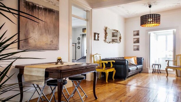 Uma sala de estar com piso de madeira, um sofá de couro preto, uma mesa de jantar com cadeiras e um grande quadro abstrato na parede.