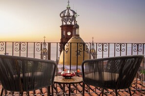 Uma varanda com duas cadeiras de vime, uma pequena mesa com uma vela e vista para uma torre com relógio.