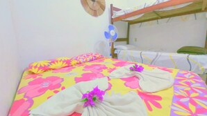 Quarto de hotel com cama coberta por um edredom estampado com flores, um ventilador e uma beliche ao fundo.