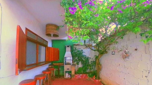 Um espaço exterior estreito com parede de tijolos, uma pequena árvore com flores roxas, uma chaminé e chão vermelho.