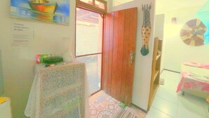 Um quarto com porta de madeira, mesa estampada, cama com colcha rosa e branca, decoração de girafa e uma pintura de barco.
