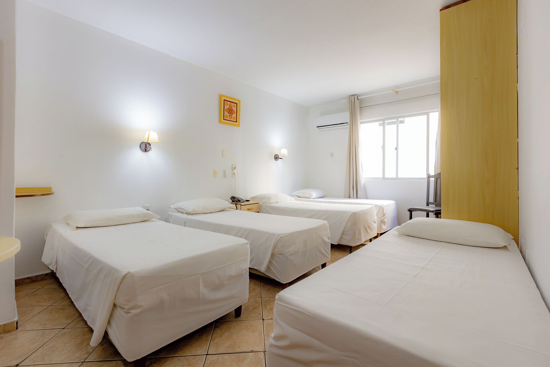 Quarto de hotel com três camas de solteiro, uma mesa de cabeceira com abajur, uma cadeira e uma janela com cortinas.