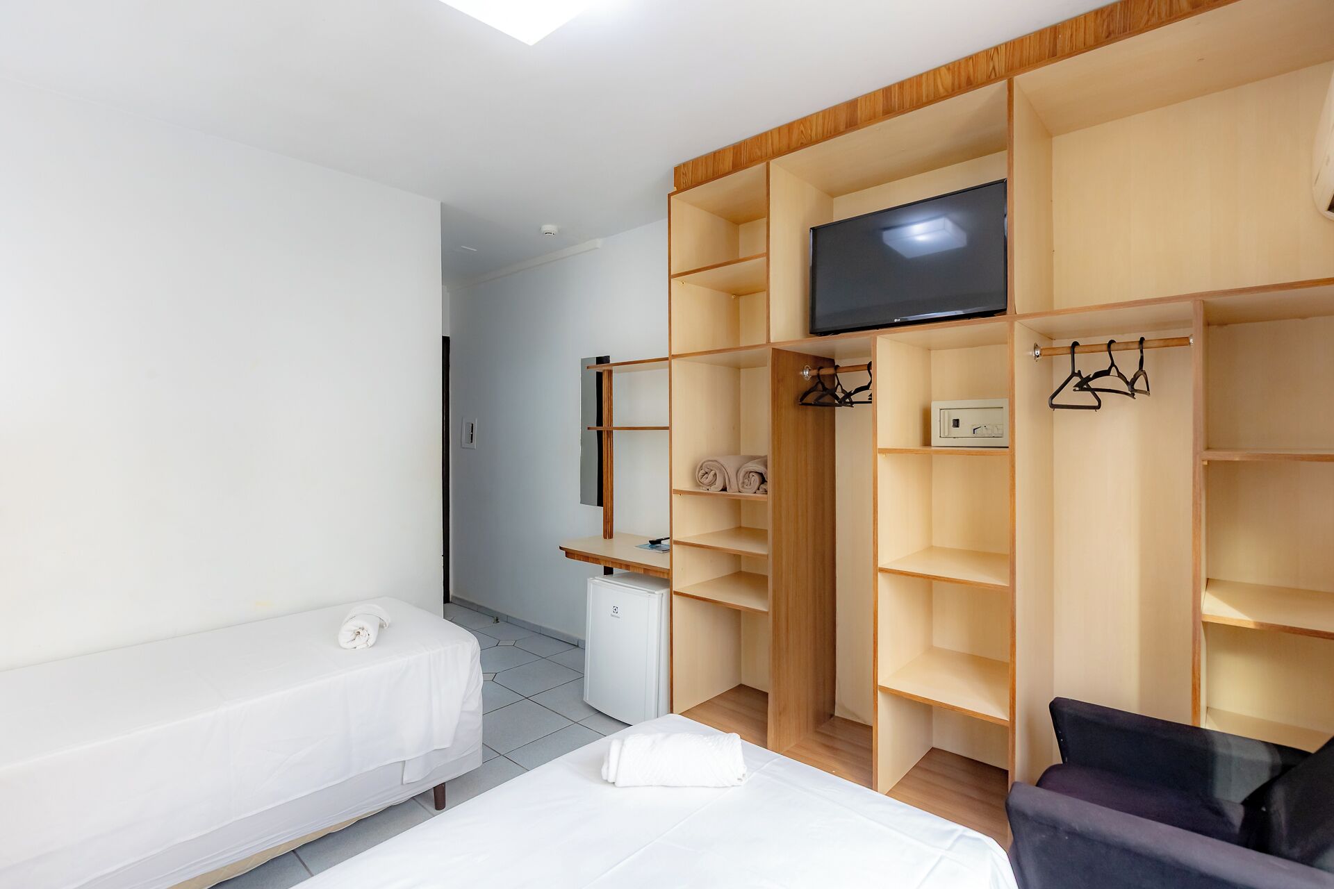 Quarto de hotel com cama, televisão presa na parede, guarda-roupa com várias prateleiras e pequena mesa com cadeira.