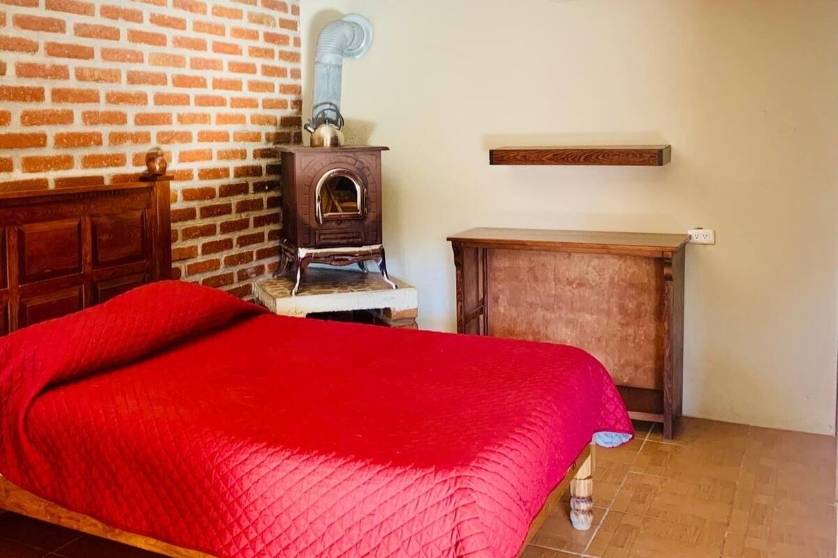 Quarto com parede de tijolos, cama de madeira com manta vermelha, fogão pequeno e unidade de ar condicionado branca.