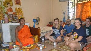 Quarto com cama, janela com cortinas, estátua de Buda e um grupo de pessoas sentadas no chão.